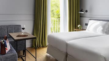 Quarto de hotel com cama, uma mesinha com um copo de vinho, uma cadeira e vista para o verde lá fora.