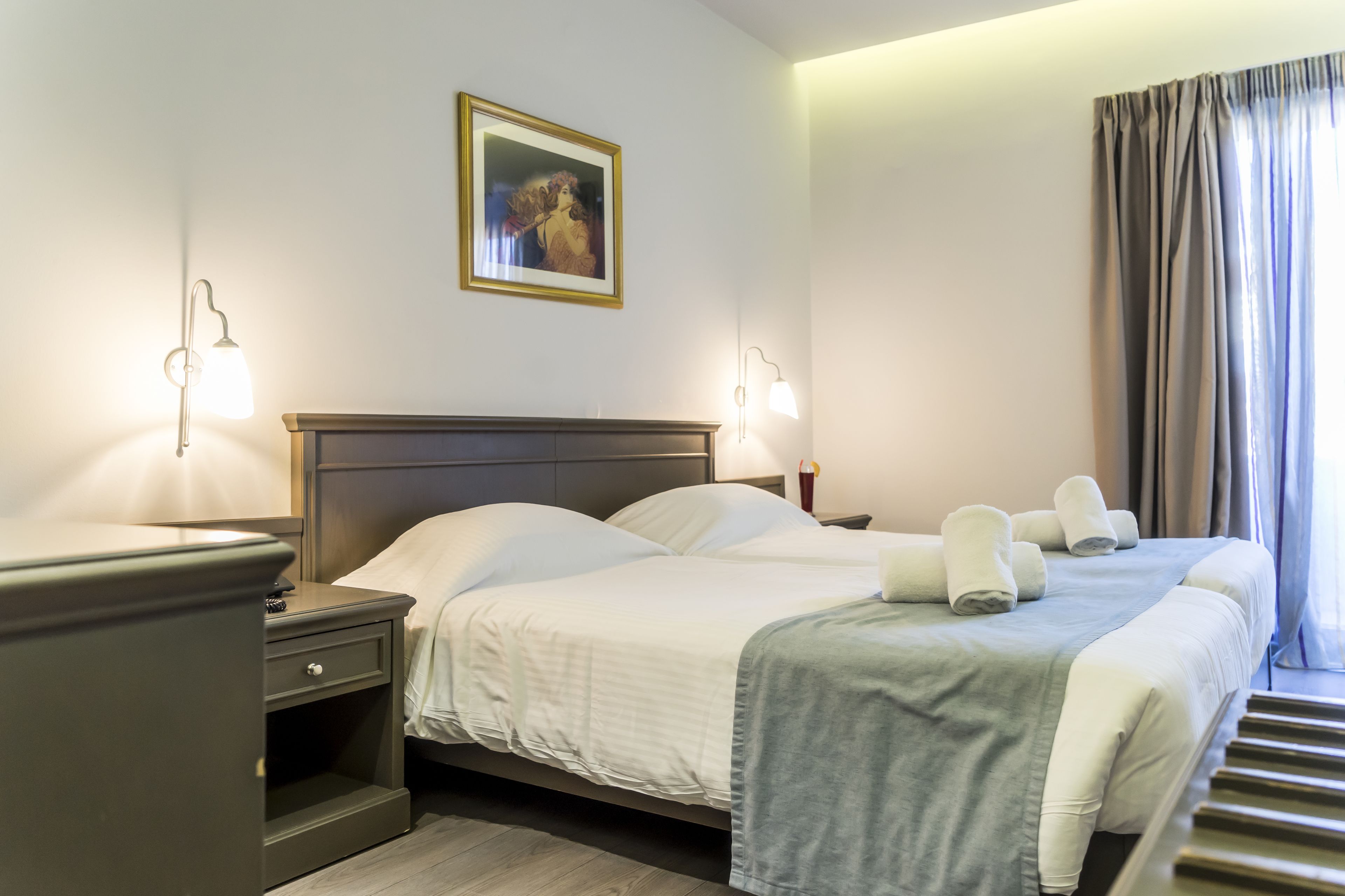 Quarto de hotel com uma cama, mesas de cabeceira, um criado-mudo, um quadro na parede e uma janela com cortinas.