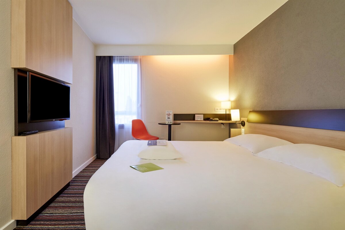 Quarto de hotel com uma cama grande, televisão de tela plana na parede, mesinha com abajur, uma cadeira vermelha e uma janela com cortinas.
