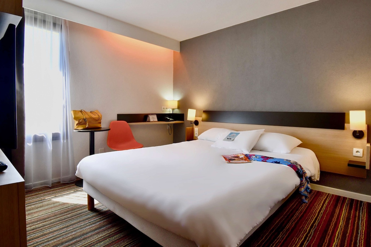 Quarto de hotel com uma cama grande, uma escrivaninha, uma cadeira e uma mesinha de cabeceira com abajur.