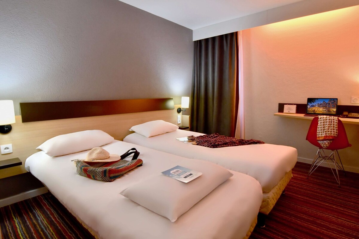 Quarto de hotel com duas camas, uma escrivaninha com um laptop, uma pequena mesa com uma cadeira vermelha e uma janela com cortinas.