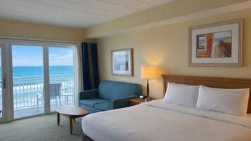 Quarto de hotel com cama, escrivaninha, cadeira, luminária, quadro e vista do oceano através de uma porta deslizante.