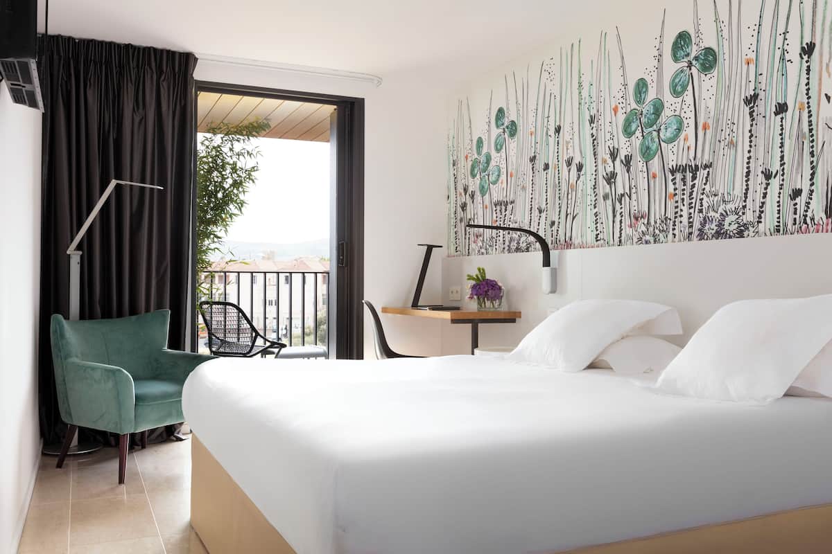 Quarto de hotel com cama, escrivaninha, cadeira e uma janela grande.