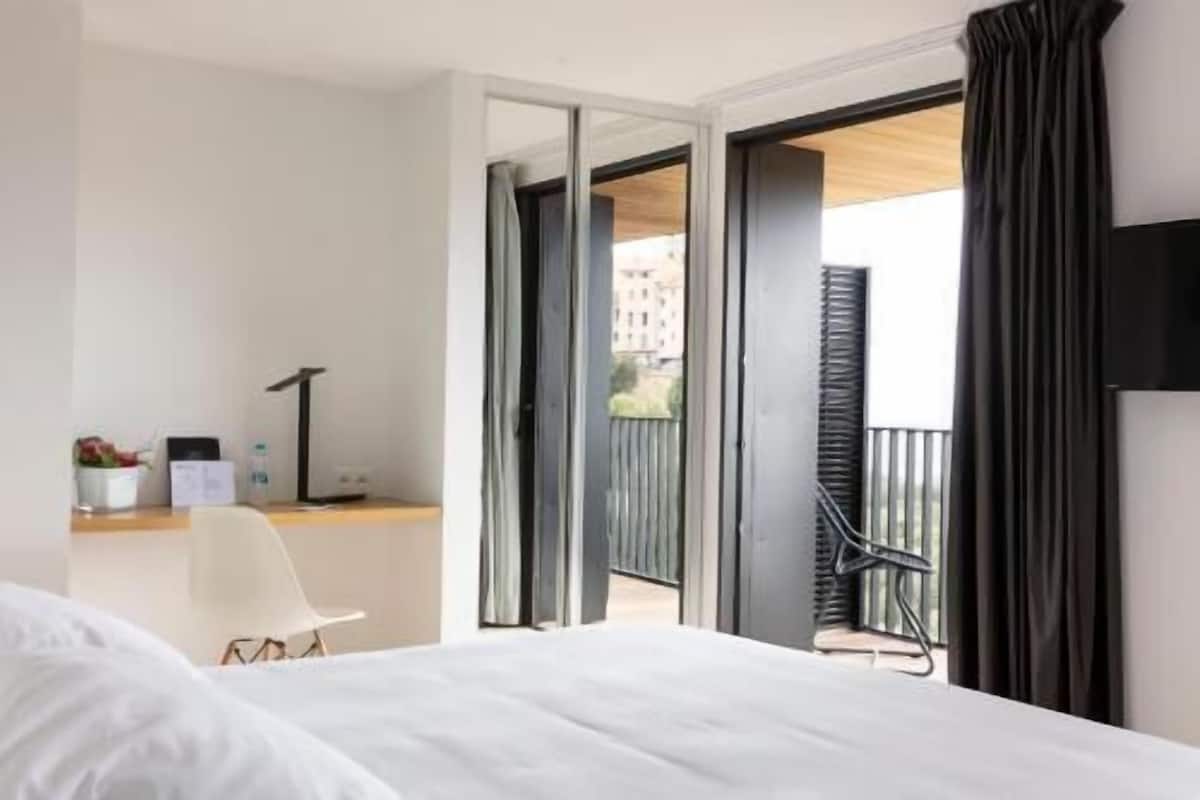 Quarto de hotel com cama, uma cadeira, escrivaninha com luminária e varanda com guarda-corpo.