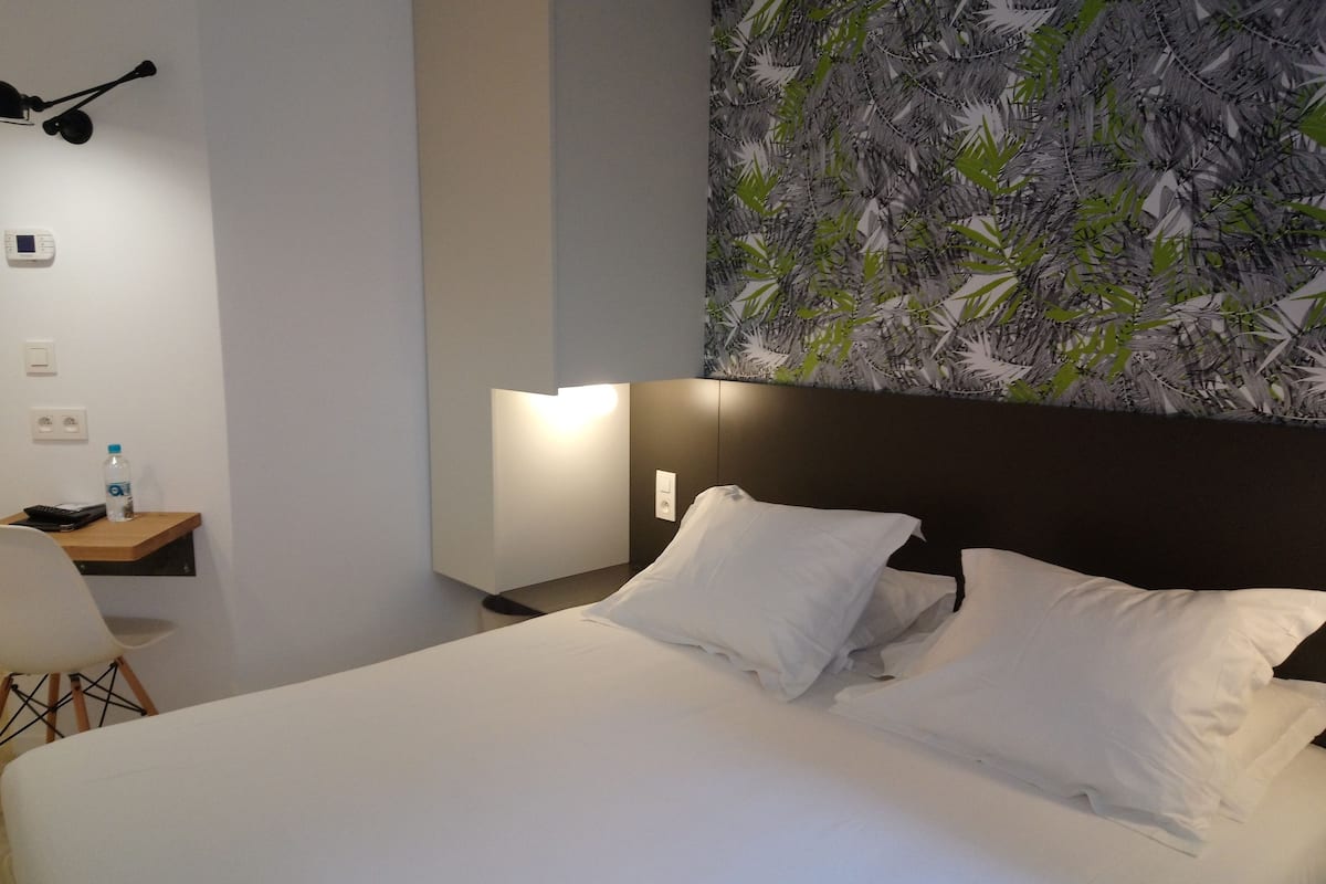 Quarto de hotel com cama, secretária com cadeira, luminária na parede e termostato.