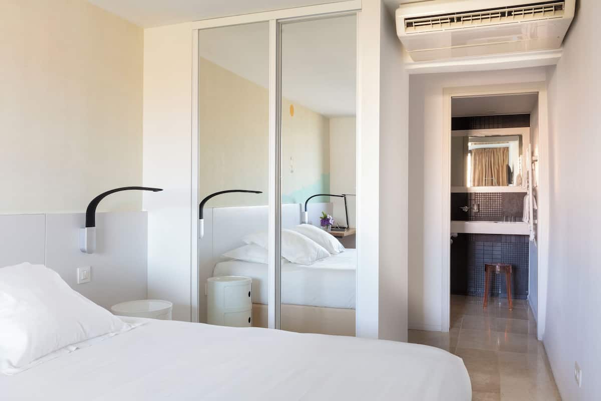 Quarto de hotel com cama, espelho, mesa pequena, mesa de cabeceira, ar condicionado fixado na parede e banheiro visível através de uma porta aberta.