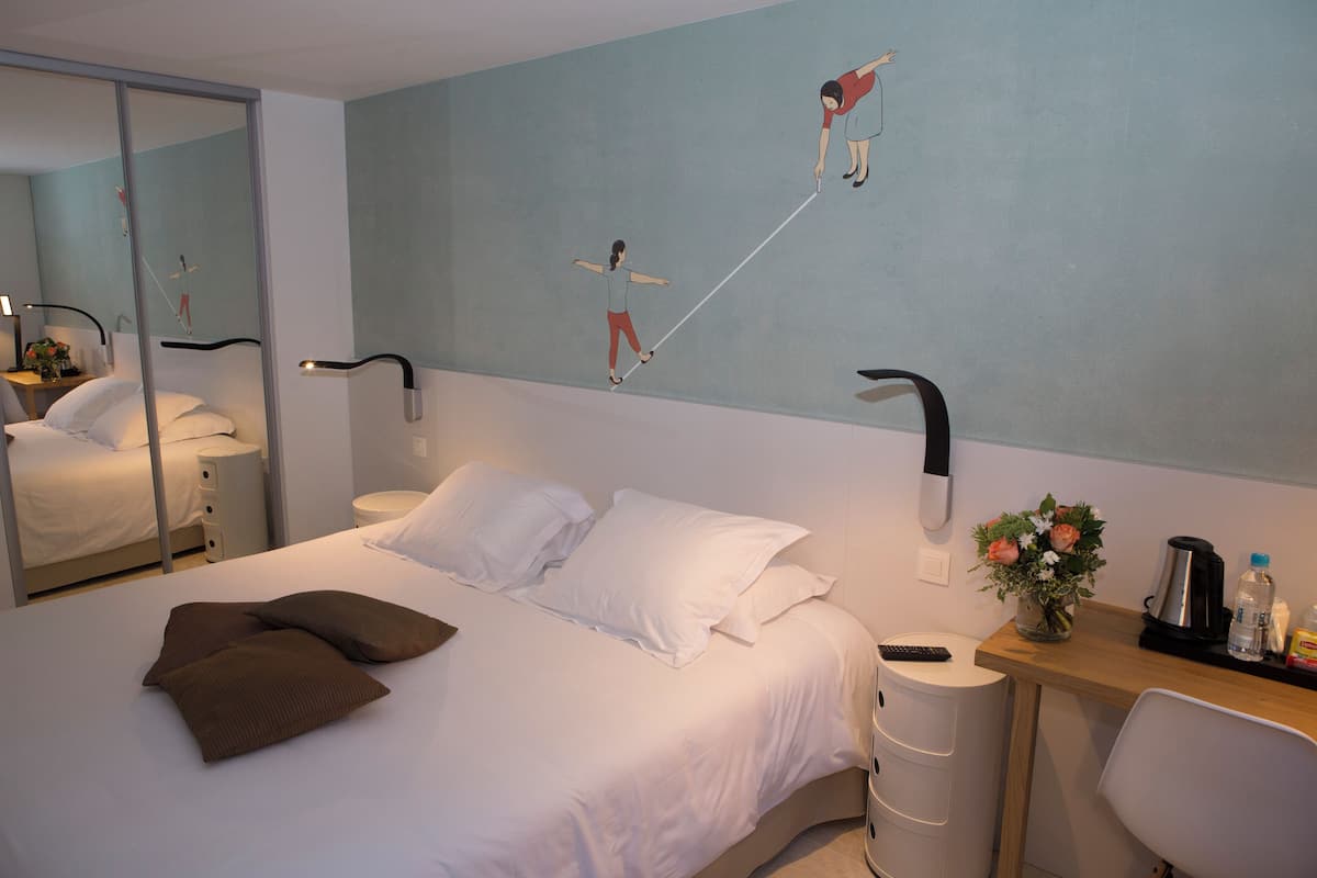 Quarto de hotel com cama, mesas de cabeceira, escrivaninha com cadeira, espelho e um quadro na parede.