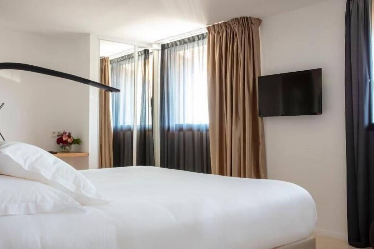 Quarto de hotel com cama, televisão presa na parede e janela com cortinas.