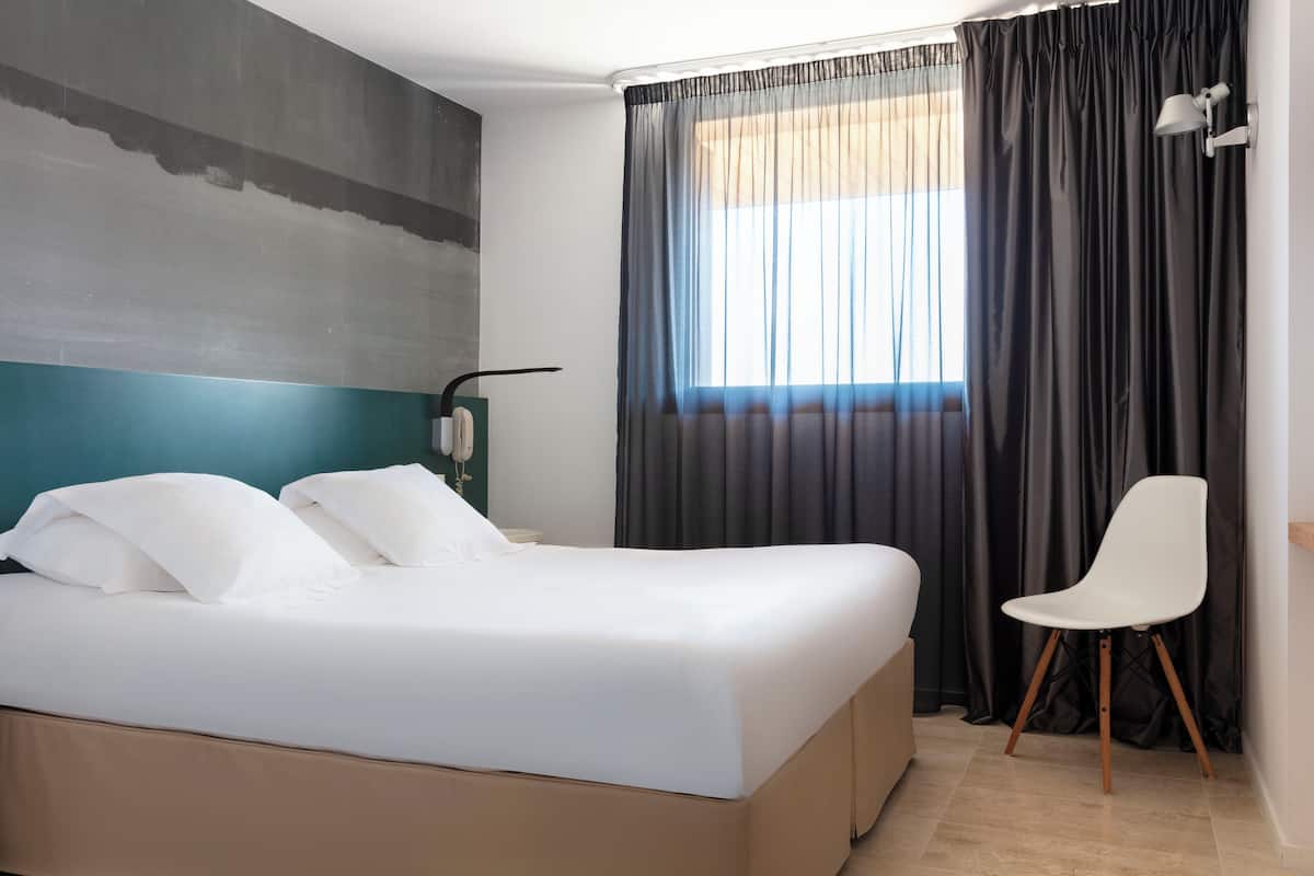 Quarto de hotel moderno com uma cama, uma poltrona, uma janela com cortinas e uma luminária na parede.