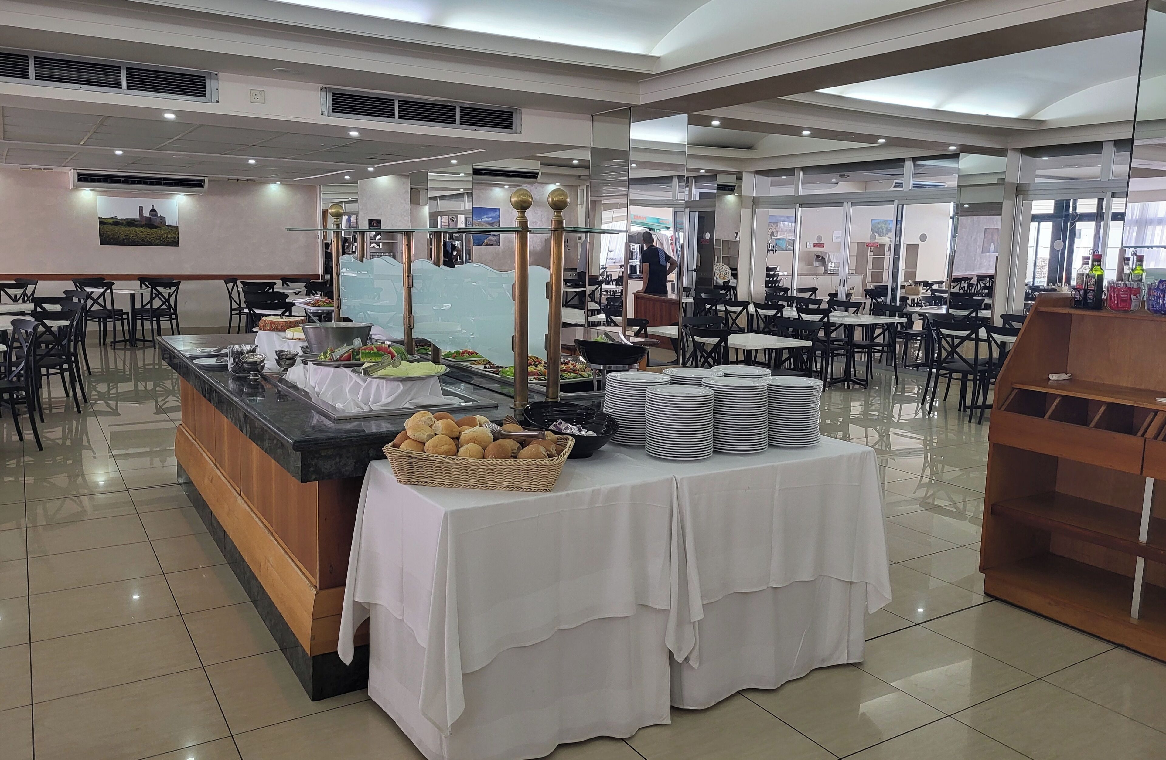 Disposição de buffet com diversos tipos de comida, um balcão com cesta de pão e pilhas de pratos bem organizadas.