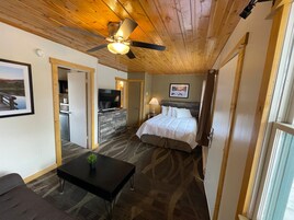Quarto com cama, escrivaninha, ventilador de teto, televisão e porta para o banheiro.