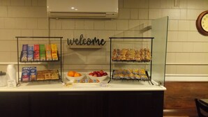 Uma área de buffet com um letreiro que diz 'welcome', lanches, frutas e bebidas.
