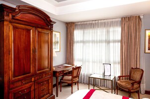Quarto de hotel com um armário de madeira grande, uma escrivaninha com cadeira, uma mesinha com cadeira e uma janela grande com cortinas.