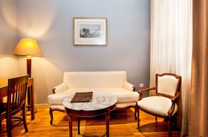 Quarto com sofá branco, cadeira de madeira, mesa redonda pequena com um livro e um copo, e uma luminária à esquerda.