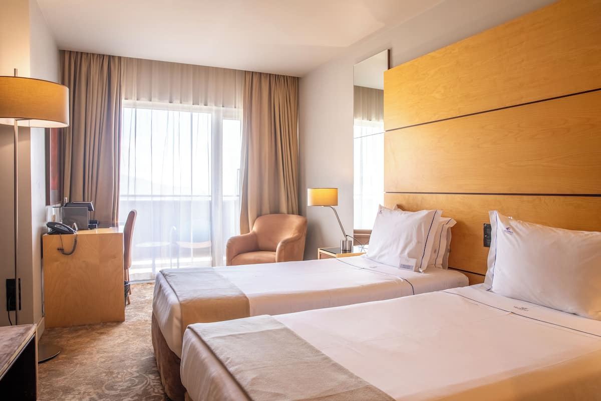 Quarto de hotel com duas camas, uma mesa de trabalho com luminária, uma cadeira e uma janela grande com cortinas.