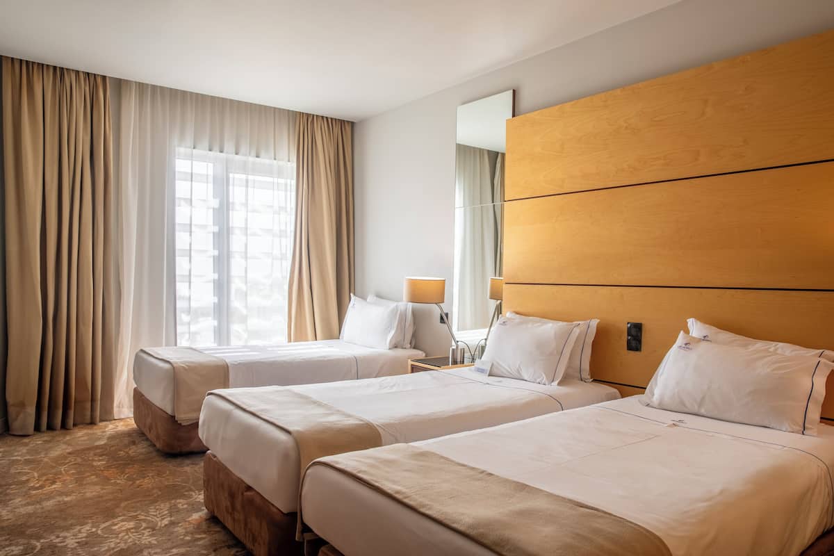 Quarto de hotel com duas camas, uma cabeceira de madeira grande e uma janela com cortinas.