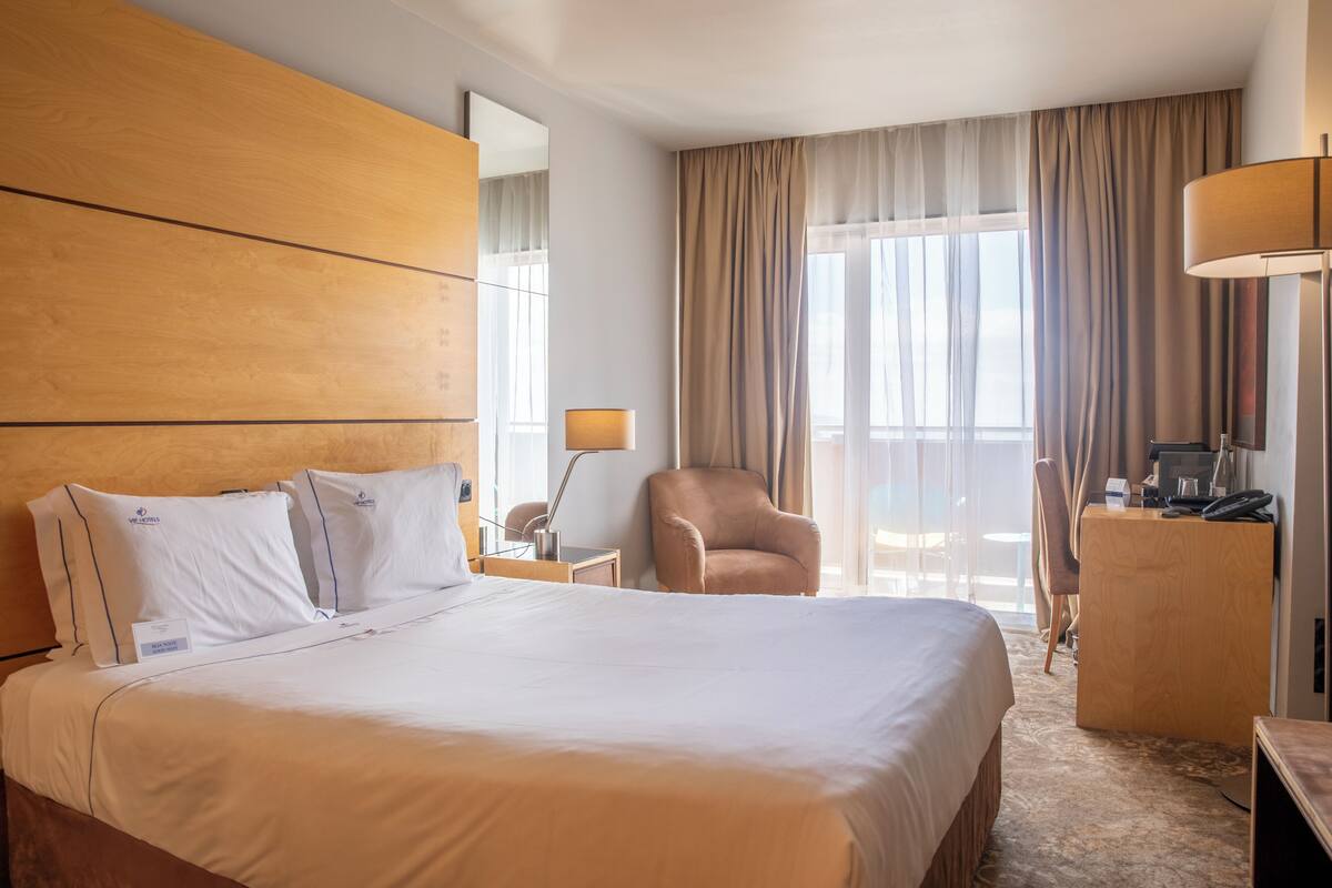 Quarto de hotel com uma cama grande, uma escrivaninha com abajur, uma cadeira e uma luminária de mesa.