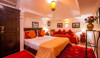 Quarto de hotel com uma cama grande, duas mesas de cabeceira, um sofá, tapete vermelho e porta de madeira.