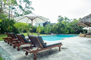 Uma área à beira da piscina com espreguiçadeiras e um grande guarda-sol, rodeada por vegetação exuberante e uma estrutura com telhado de palha.