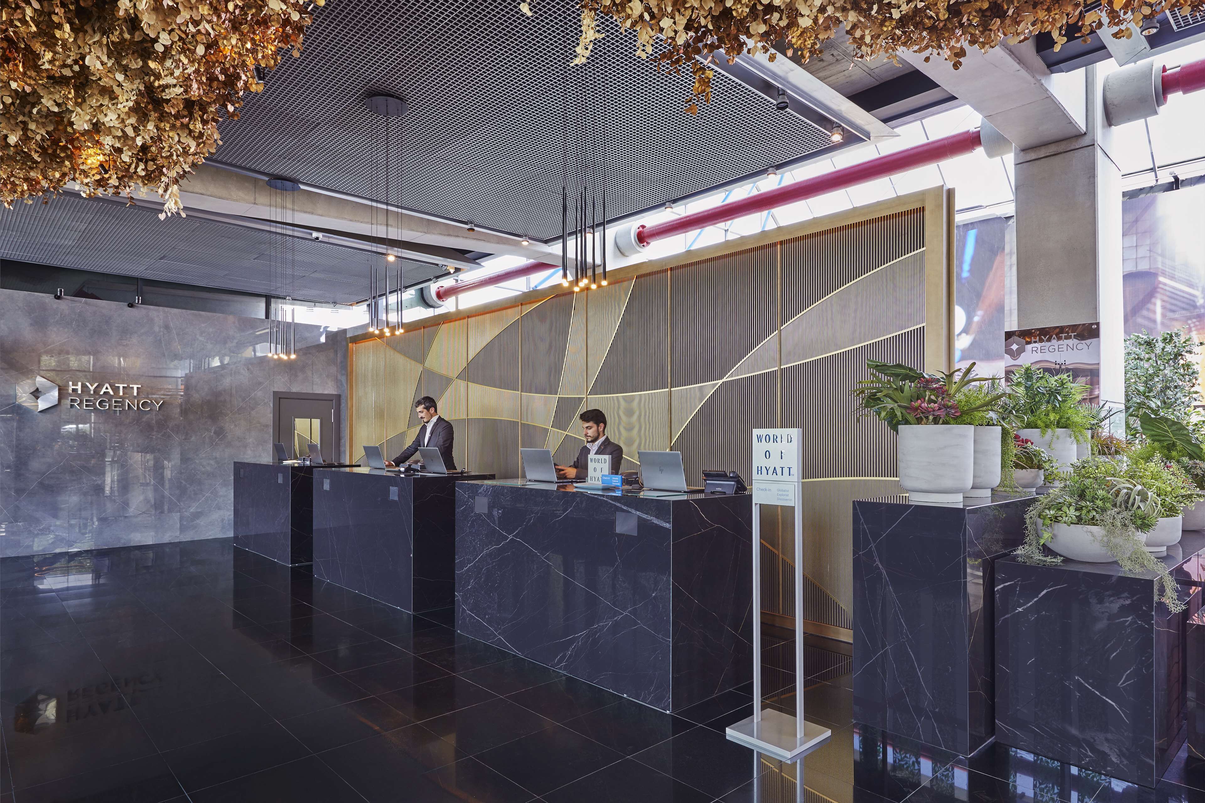 Uma área de recepção de hotel moderna com balcões de mármore, dois recepcionistas em seus postos e uma grande planta no canto.