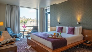 Quarto de hotel moderno com uma cama grande, uma escrivaninha com um laptop, uma cadeira e vista para a cidade através de amplas janelas.