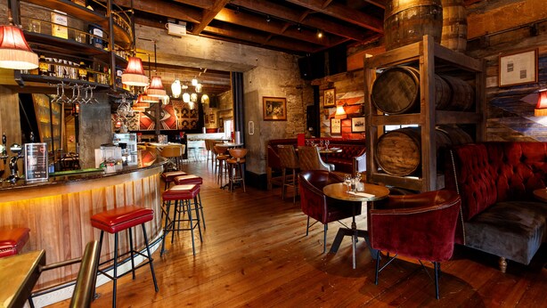 Uma área de bar rústica com chão de madeira, banquetas vermelhas e diversas garrafas e copos em prateleiras.