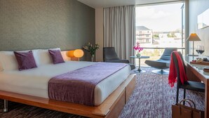 Quarto de hotel com uma cama grande, uma escrivaninha com cadeira, uma mesinha de cabeceira com abajur, e vista para prédios pela janela.