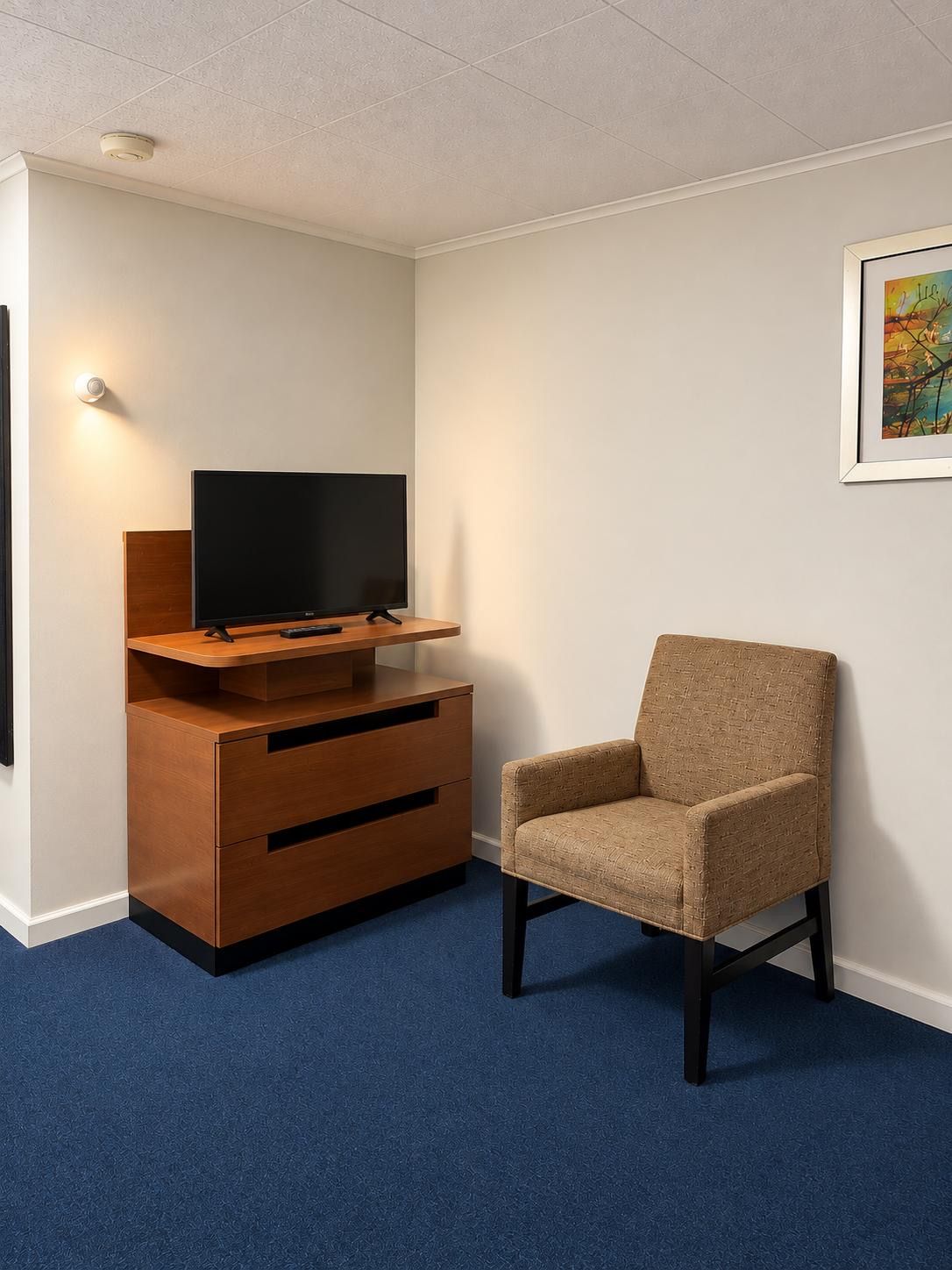 Quarto com televisão sobre um suporte de madeira, uma poltrona marrom e um quadro na parede.