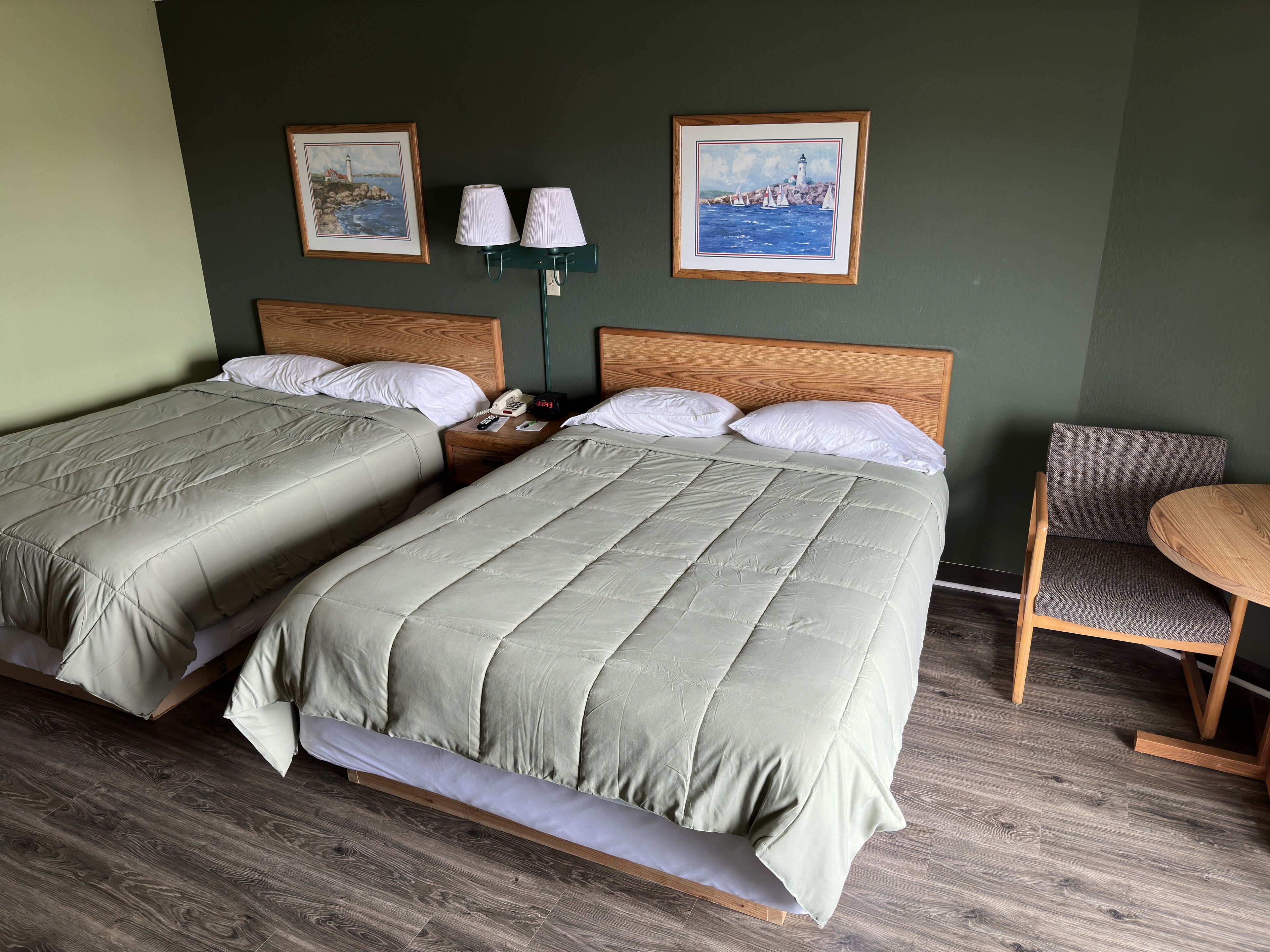 Quarto de hotel com duas camas, uma mesa de cabeceira com telefone e luminária, uma cadeira e duas quadros na parede.