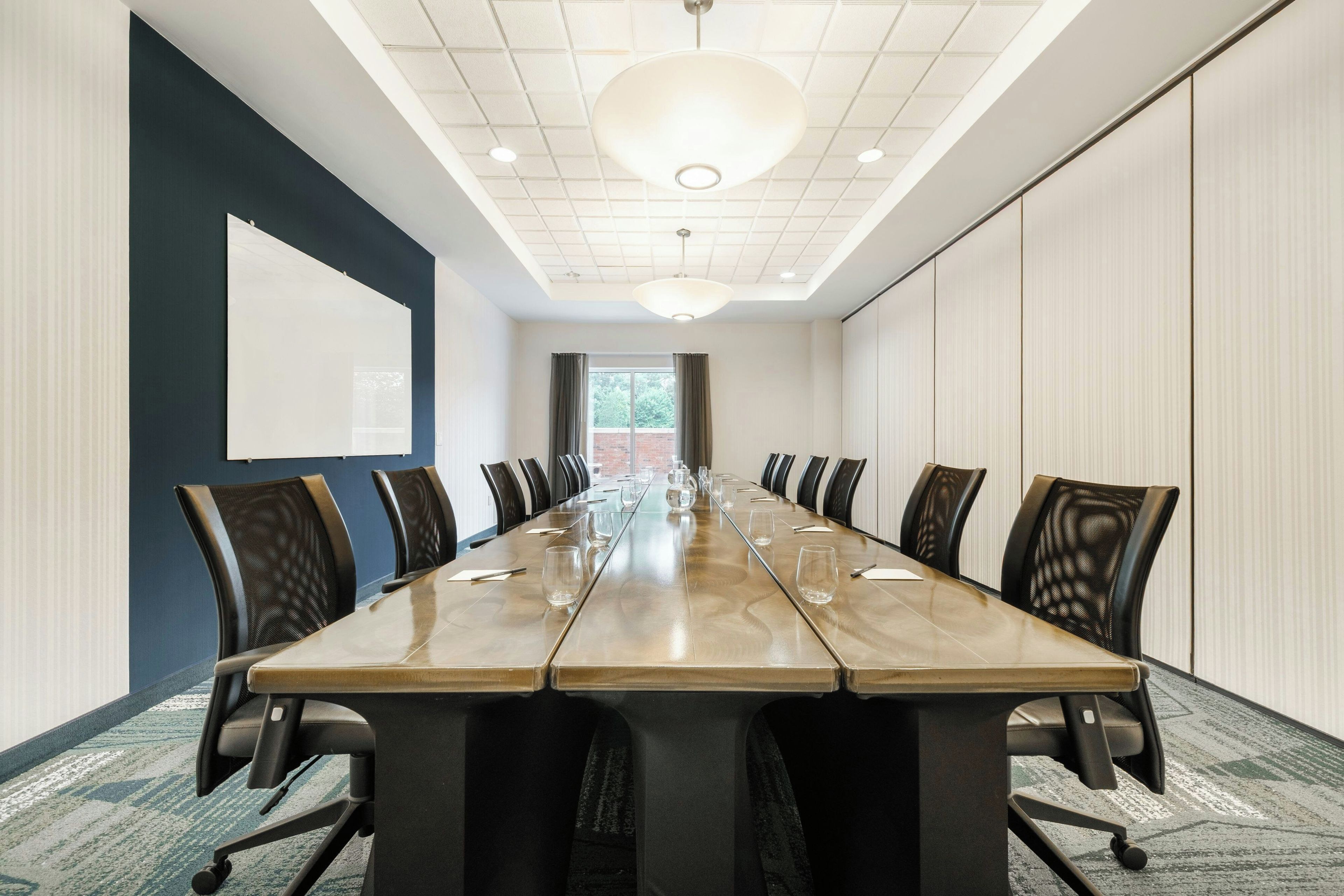 Sala de conferência com uma mesa comprida, cadeiras, tela de projetor e vista para o exterior.