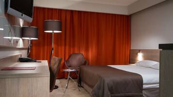 Quarto de hotel com cama, escrivaninha com telefone, cadeira, mesa pequena com abajur e televisão na parede.