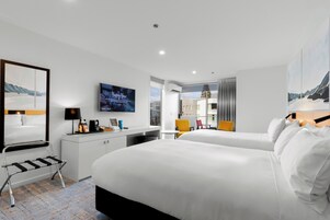 Quarto de hotel com duas camas, televisão, secretária com candeeiro e vista para a cidade.