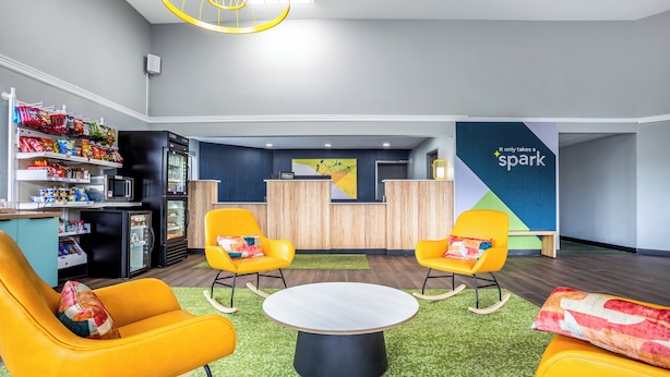 Um lobby moderno com balcão de recepção, duas cadeiras amarelas, uma mesa redonda e um bar com diversos alimentos.