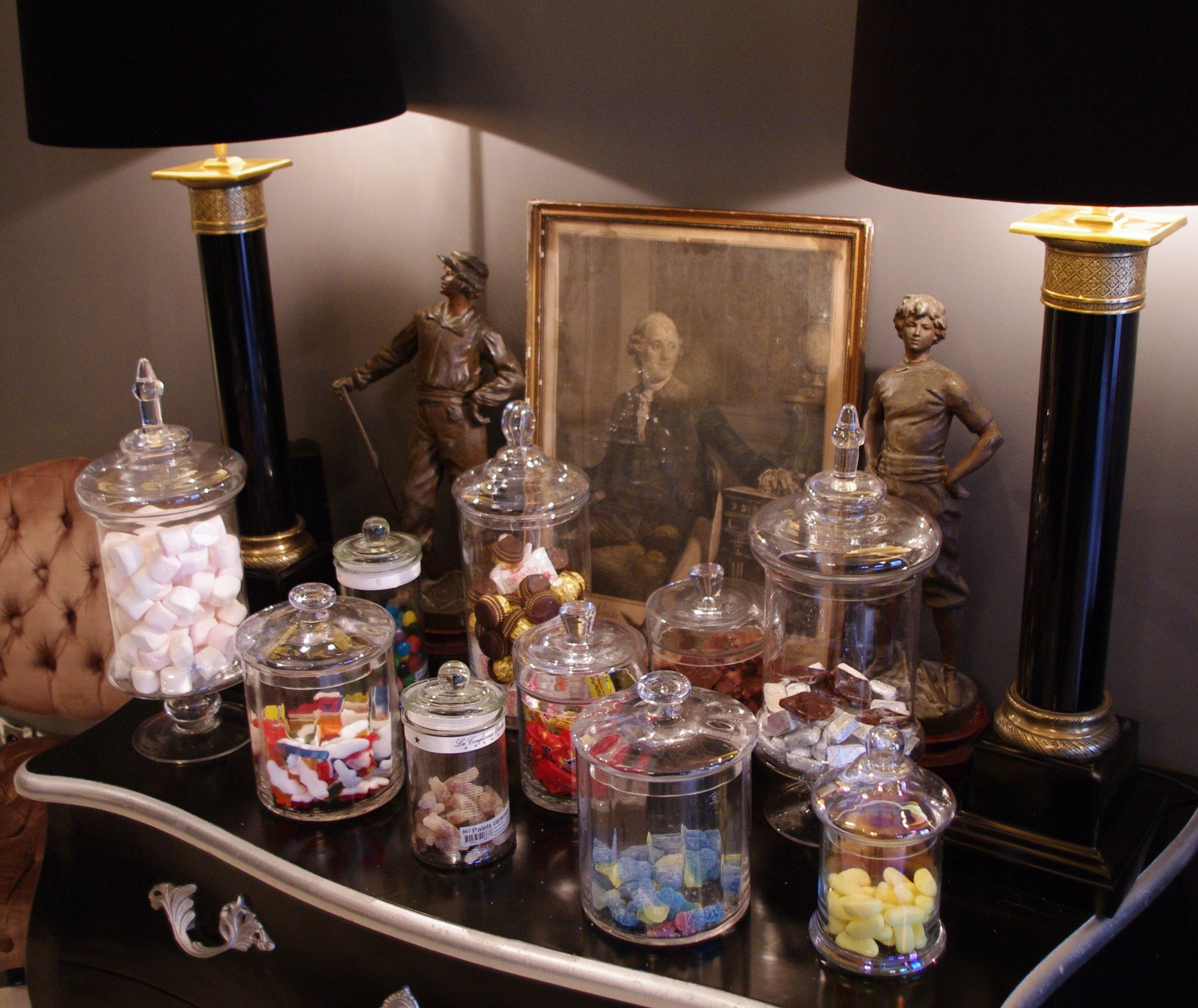 Exposição de potes de vidro cheios de diversos doces e chocolates, dispostos sobre uma mesa com uma moldura e dois objetos semelhantes a velas ao fundo.
