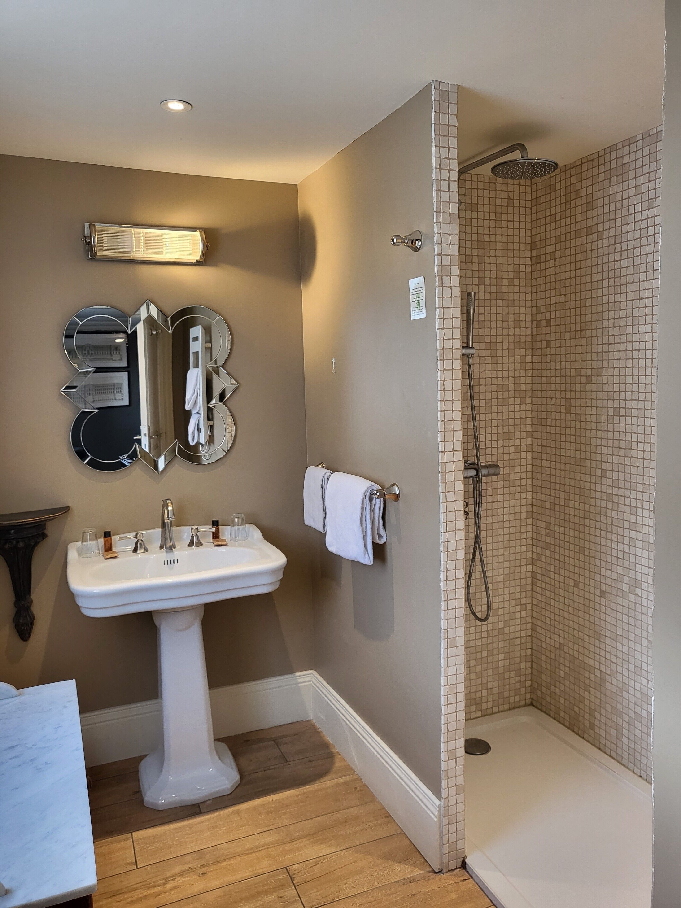 Um banheiro com pia branca de pedestal, espelho com desenhos geométricos, porta-toalhas e chuveiro com azulejos de mosaico.