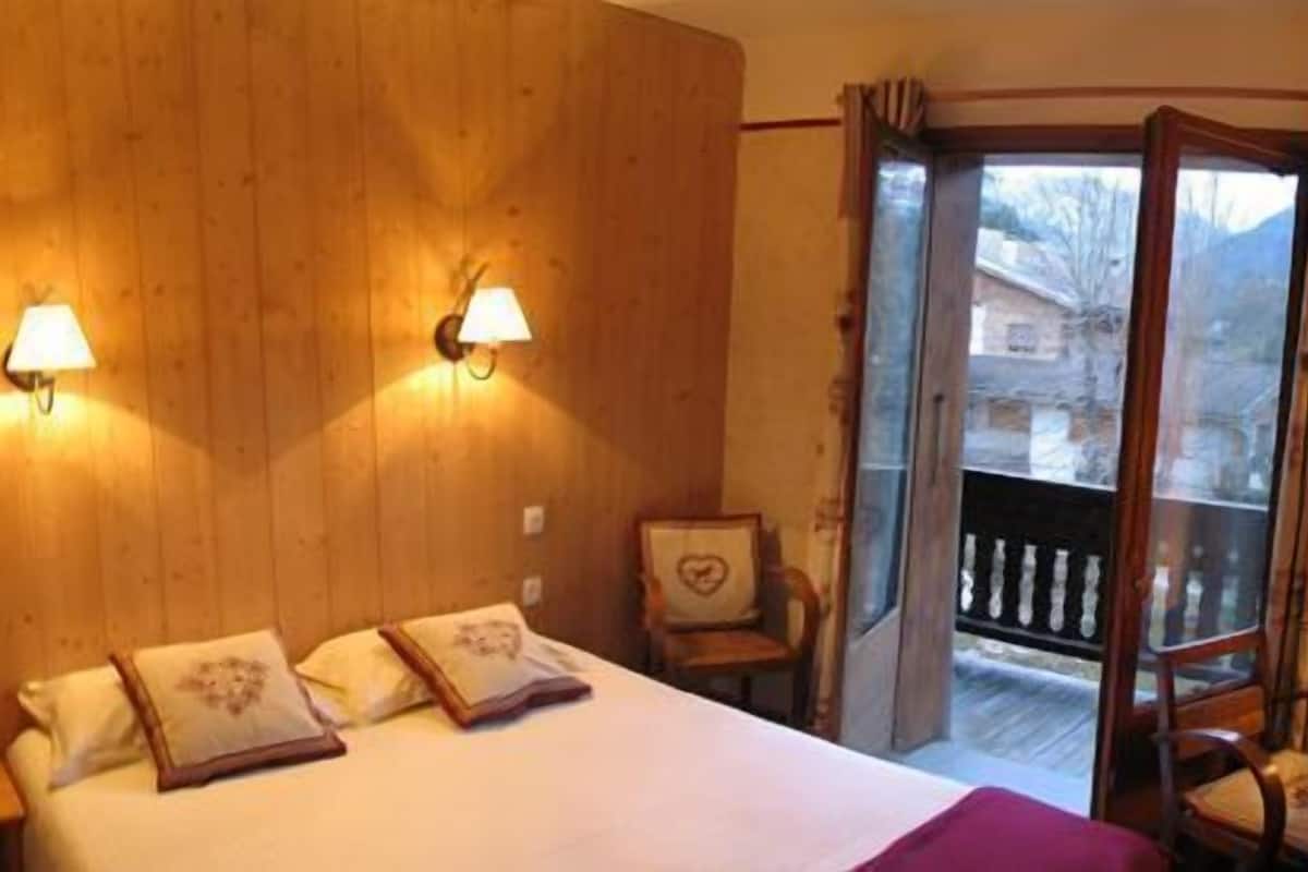 Quarto de hotel com parede de painéis de madeira, uma cama com almofadas, uma cadeira, uma mesa com abajur e uma varanda com vista.