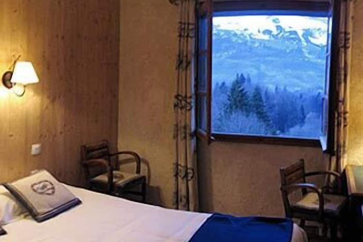 Quarto com cama, duas cadeiras, janela com vista para uma paisagem nevada e luminária fixada na parede.