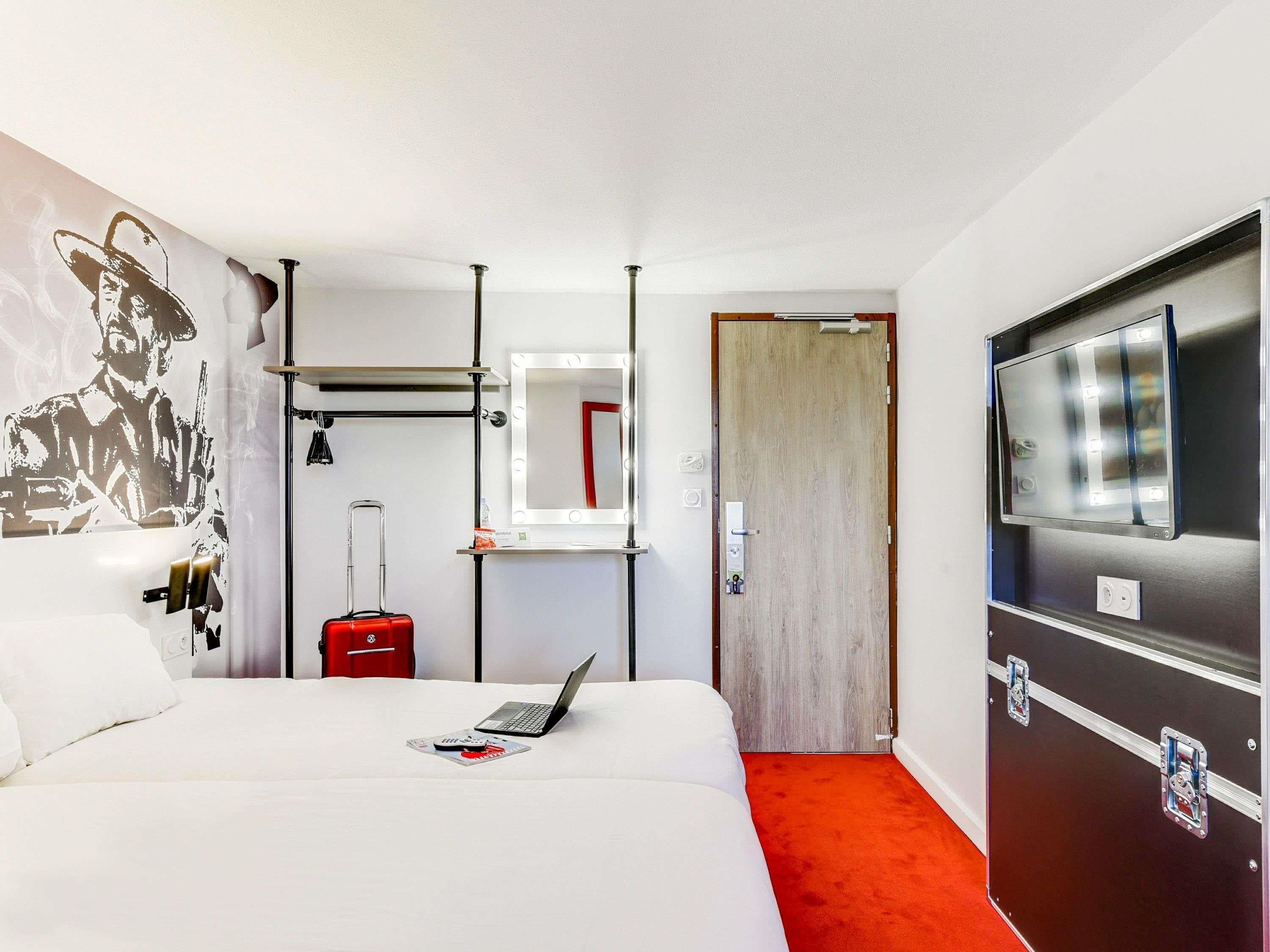 Quarto de hotel com uma cama, uma escrivaninha com um laptop, uma mala, uma televisão na parede e uma porta com espelho.