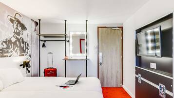 Quarto de hotel com uma cama, uma escrivaninha com um laptop, uma mala, uma televisão na parede e uma porta com espelho.