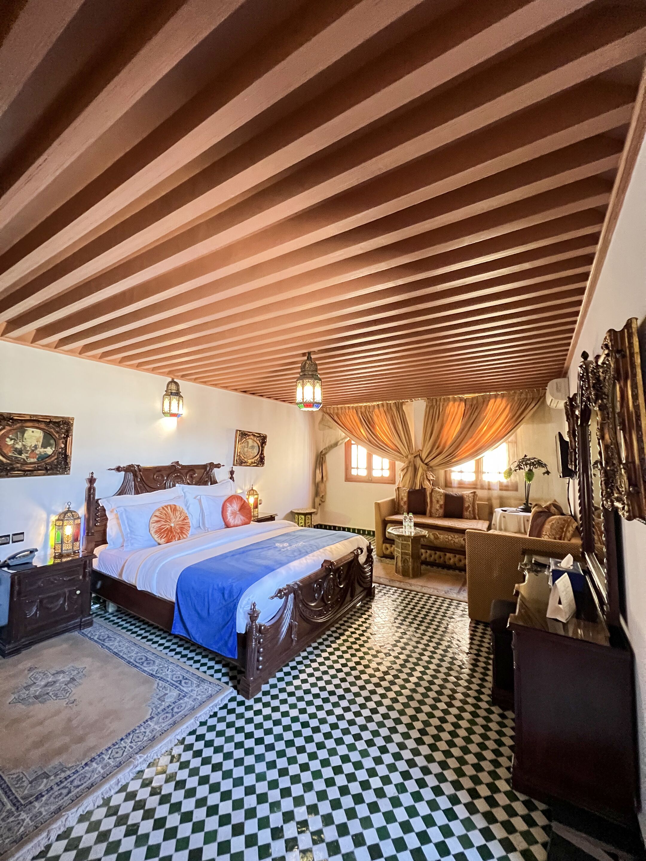 Um quarto com piso xadrez, cama com colcha azul, teto de madeira e um lustre.
