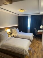 Quarto de hotel com duas camas, uma escrivaninha, uma cadeira e um ventilador de teto.
