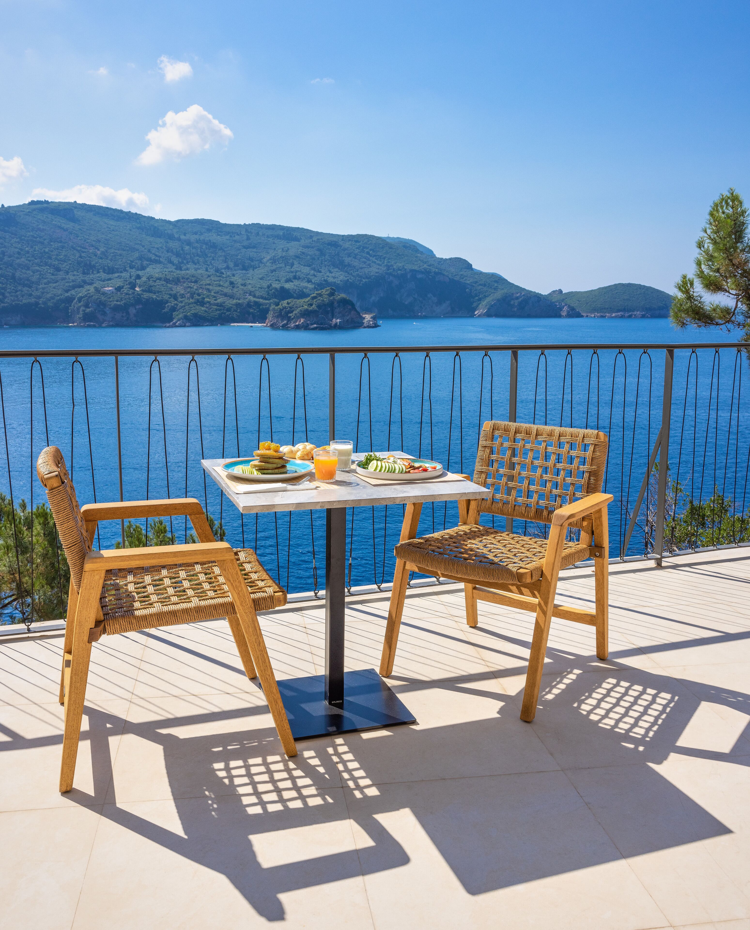Uma mesa com duas cadeiras de vime e uma refeição num terraço com vista para um lago e montanhas.