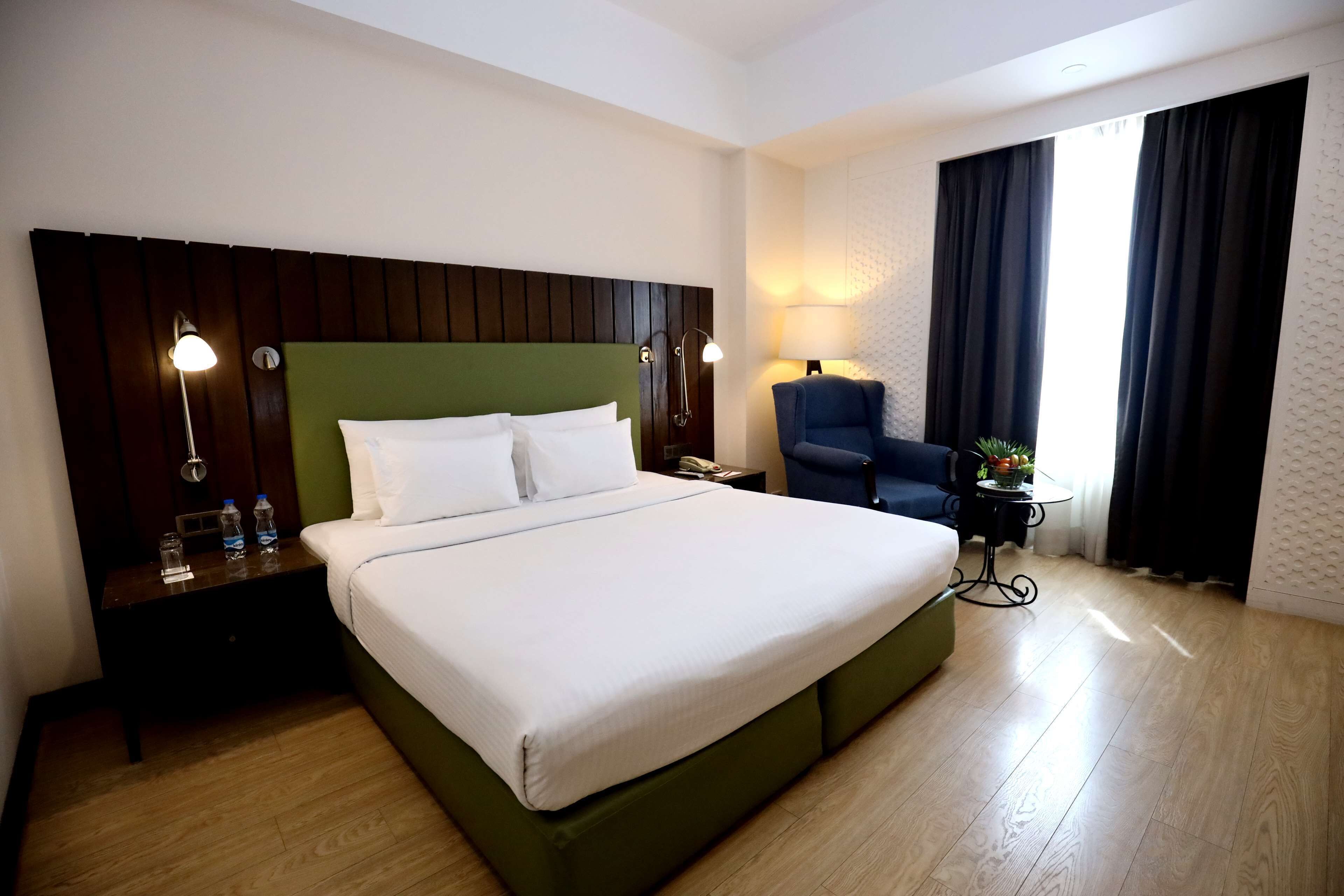 Quarto de hotel com uma cama grande, duas mesas de cabeceira, uma cadeira, uma pequena mesa com uma planta e uma janela com cortinas.