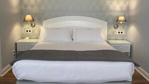 Uma cama grande com cabeceira cinza, duas mesas de cabeceira com abajures e papel de parede estampado.