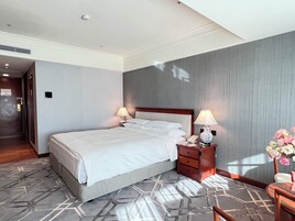 Quarto de hotel com uma cama grande, duas mesas de cabeceira com luminárias, um criado-mudo de madeira e um vaso de flores.