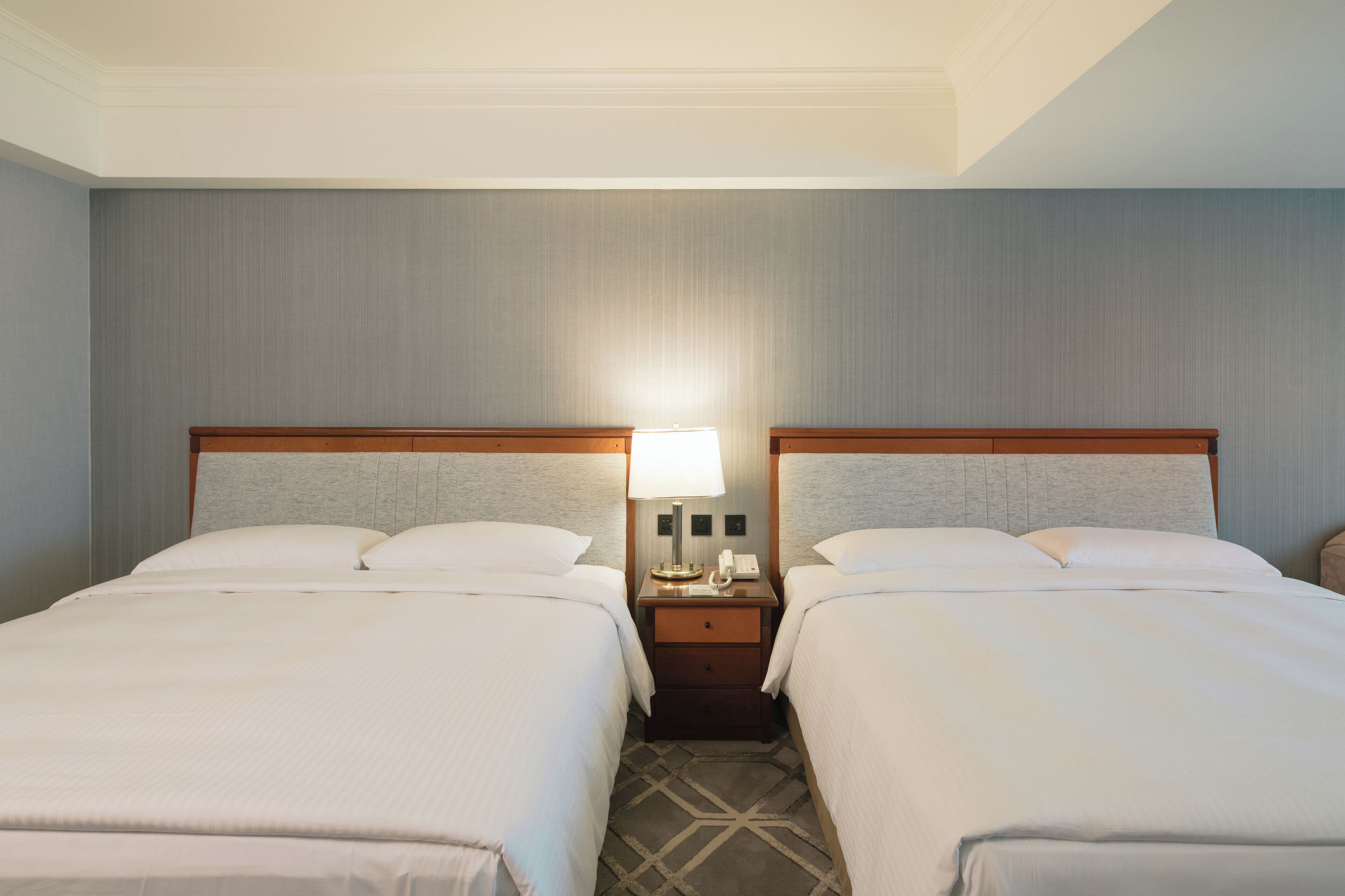 Quarto de hotel com duas camas, uma mesa de cabeceira com abajur e um telefone.