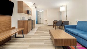 Quarto de hotel moderno com um banco de madeira, um sofá azul, uma mesa de jantar com cadeiras, micro-ondas e uma luminária.