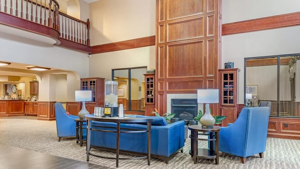 Um hall amplo com teto alto, uma escadaria imponente e uma lareira. Há poltronas estofadas em azul e uma mesa de centro.