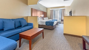 Quarto de hotel com sofá azul, mesa de centro de madeira, cama com colcha azul e escrivaninha com luminária.