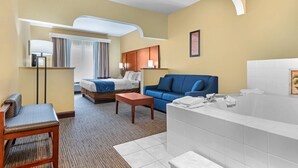 Quarto de hotel com cama, sofá, mesa pequena, escrivaninha, luminária, moldura de quadro e banheira.